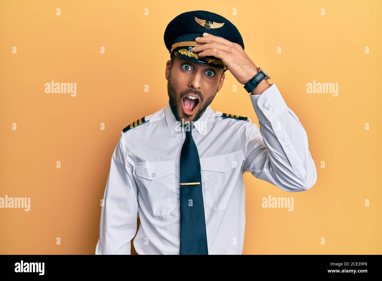 Handsome hispanic man wearing airplane pilot uniform surprised with ...