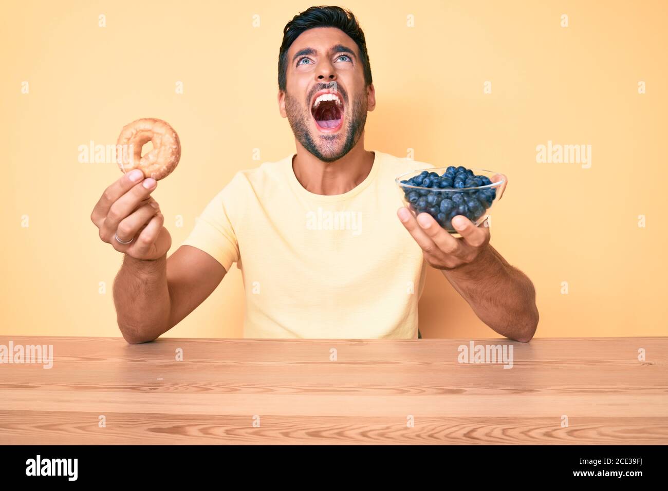 Young hispanic man eating breakfast holding donut and blueberries angry ...