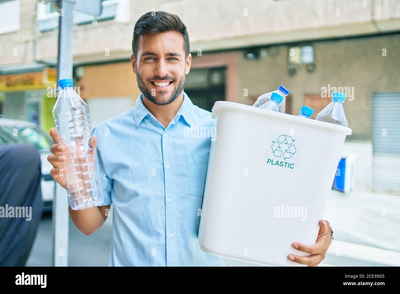 Young hispanic man smiling happy recycling. Holding full bin of plastic ...