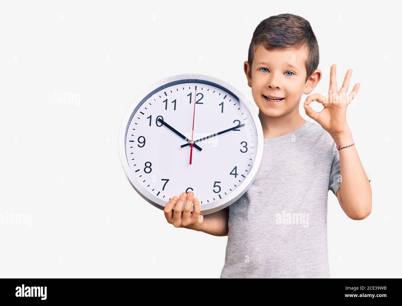 Cute blond kid holding big clock doing ok sign with fingers, smiling ...