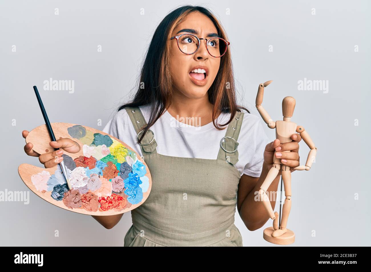 Young latin woman artist holding palette and manikin angry and mad ...