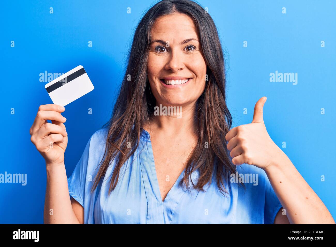 Young beautiful brunette woman holding credit card over isolated blue ...