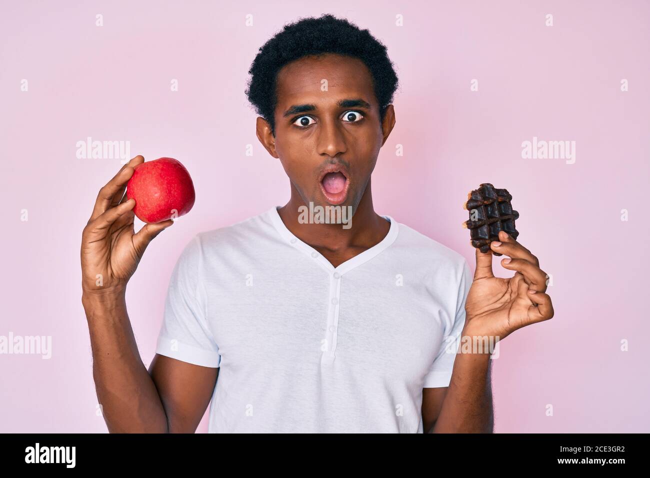 Handsome african man holding red apple and chocolate waffle afraid and ...