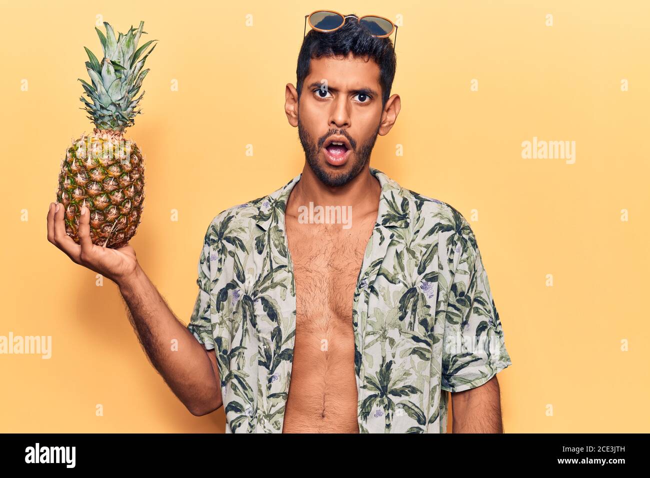 Young latin man wearing summer shirt holding pineapple scared and ...