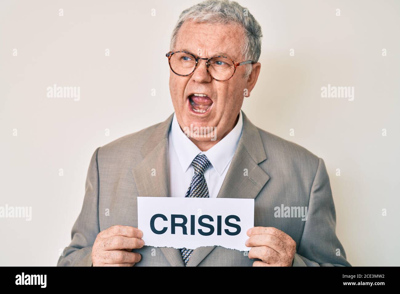 Senior grey-haired man wearing business suit holding crisis paper angry ...