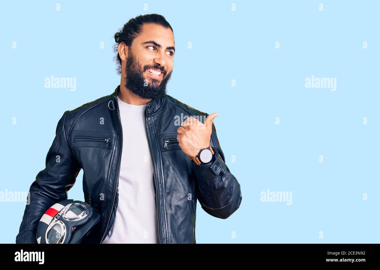 Young arab man holding motorcycle helmet pointing thumb up to the side ...
