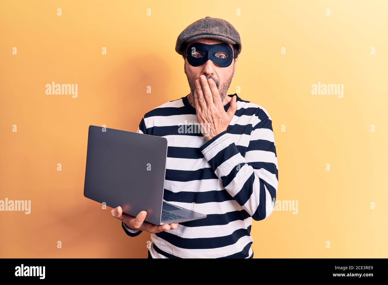 Middle age handsome burglar man wearing cap and mask using laptop over ...