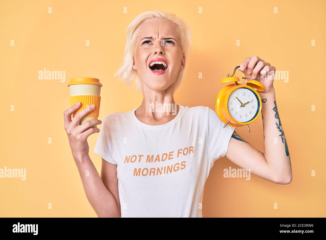 Young blonde woman with tattoo holding alarm clock and take away cup of ...