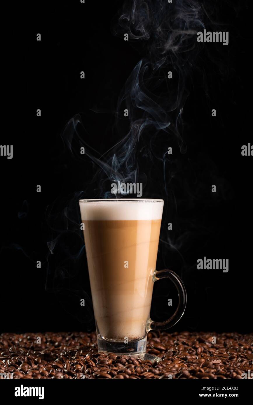 Steaming coffee latte with frothy milk in tall glass, on coffee beans ...