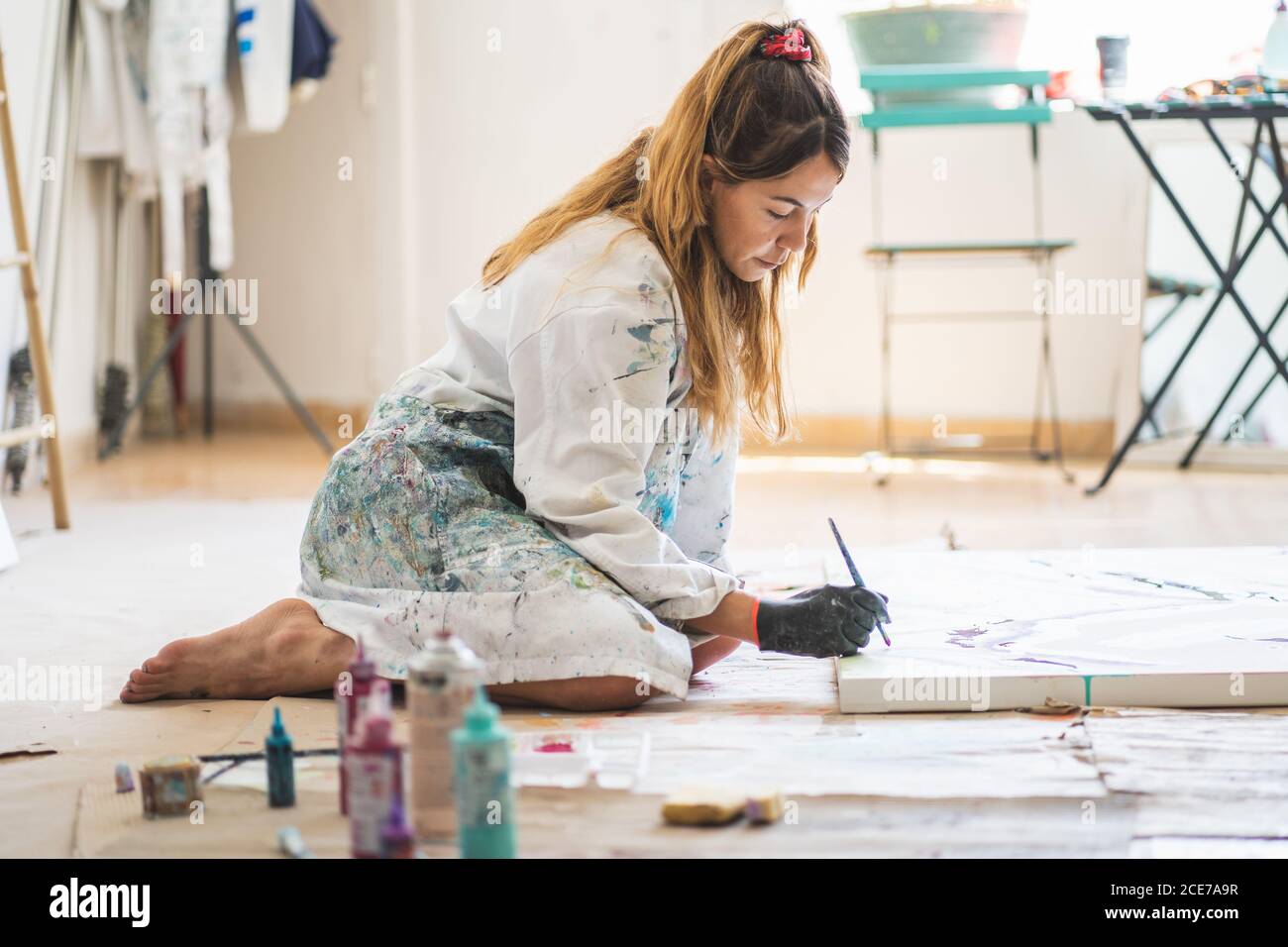 Cheerful barefoot female artist in dirty robe and gloves lying on ...