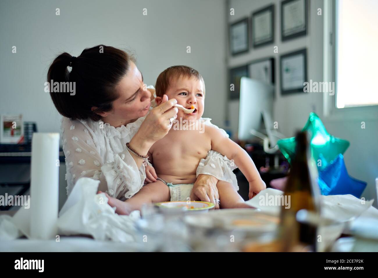 Crying little baby sitting on knees of mother and refusing food from ...