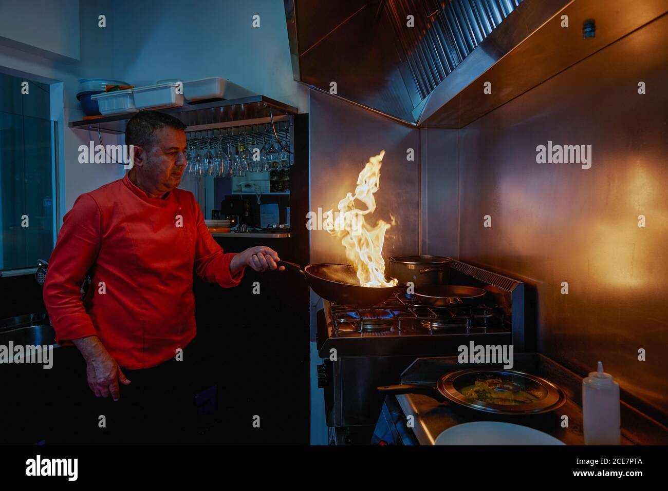 Concentrated chef in uniform holding frying pan while flambeing ...