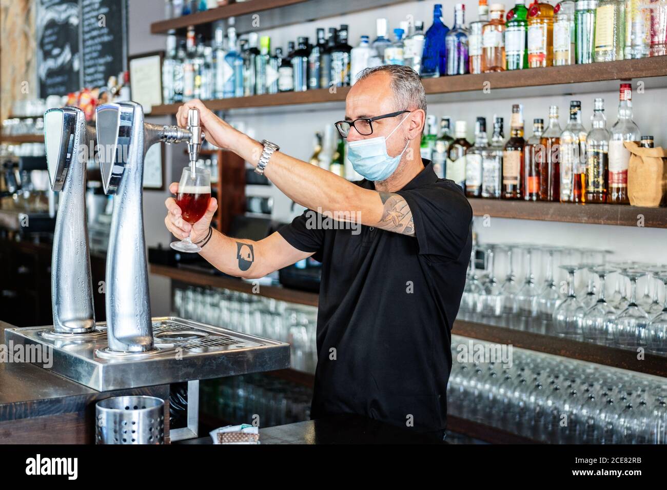 Senior ethnic male barkeeper in medical mask pouring alcohol into glass ...
