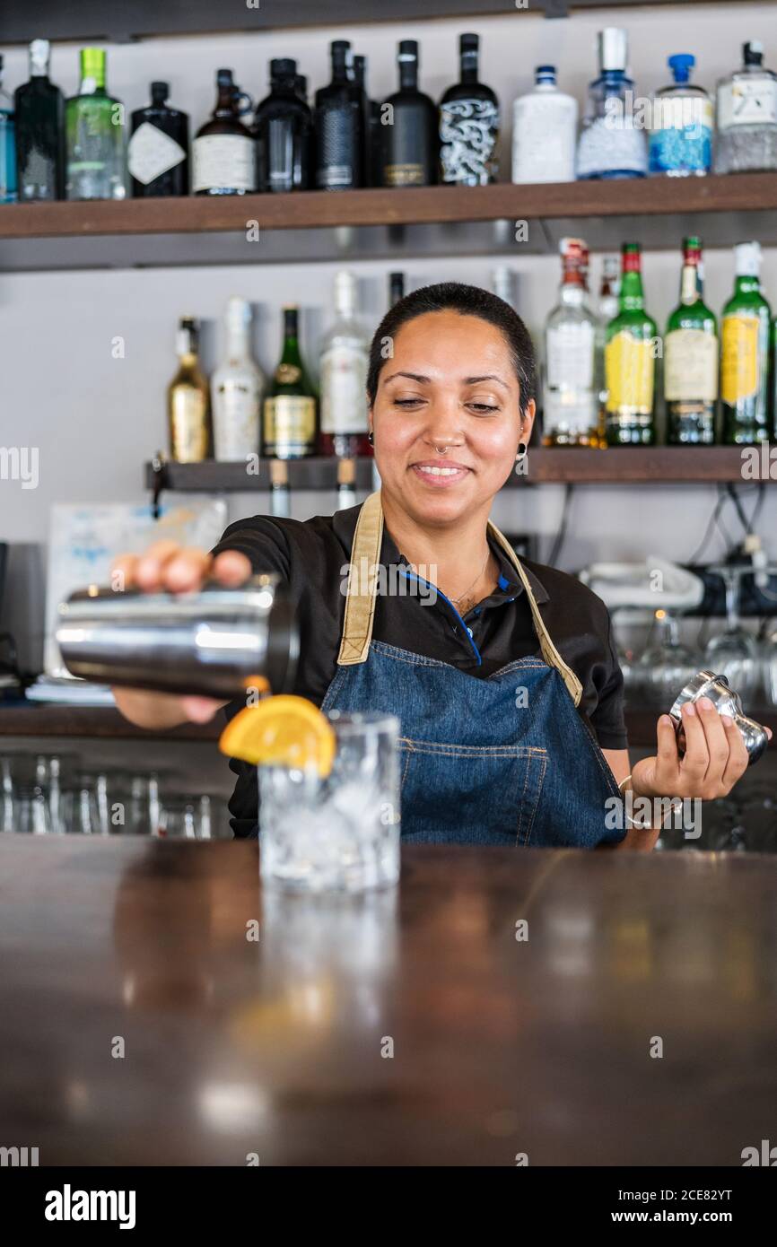 Positive focused female barkeeper in apron standing at counter making ...