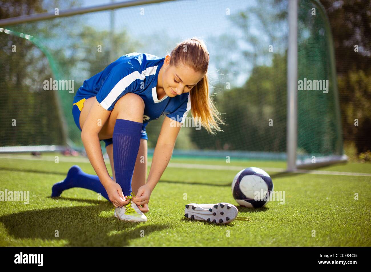 Glad female athlete putting on and tying boots near ball and goal while ...