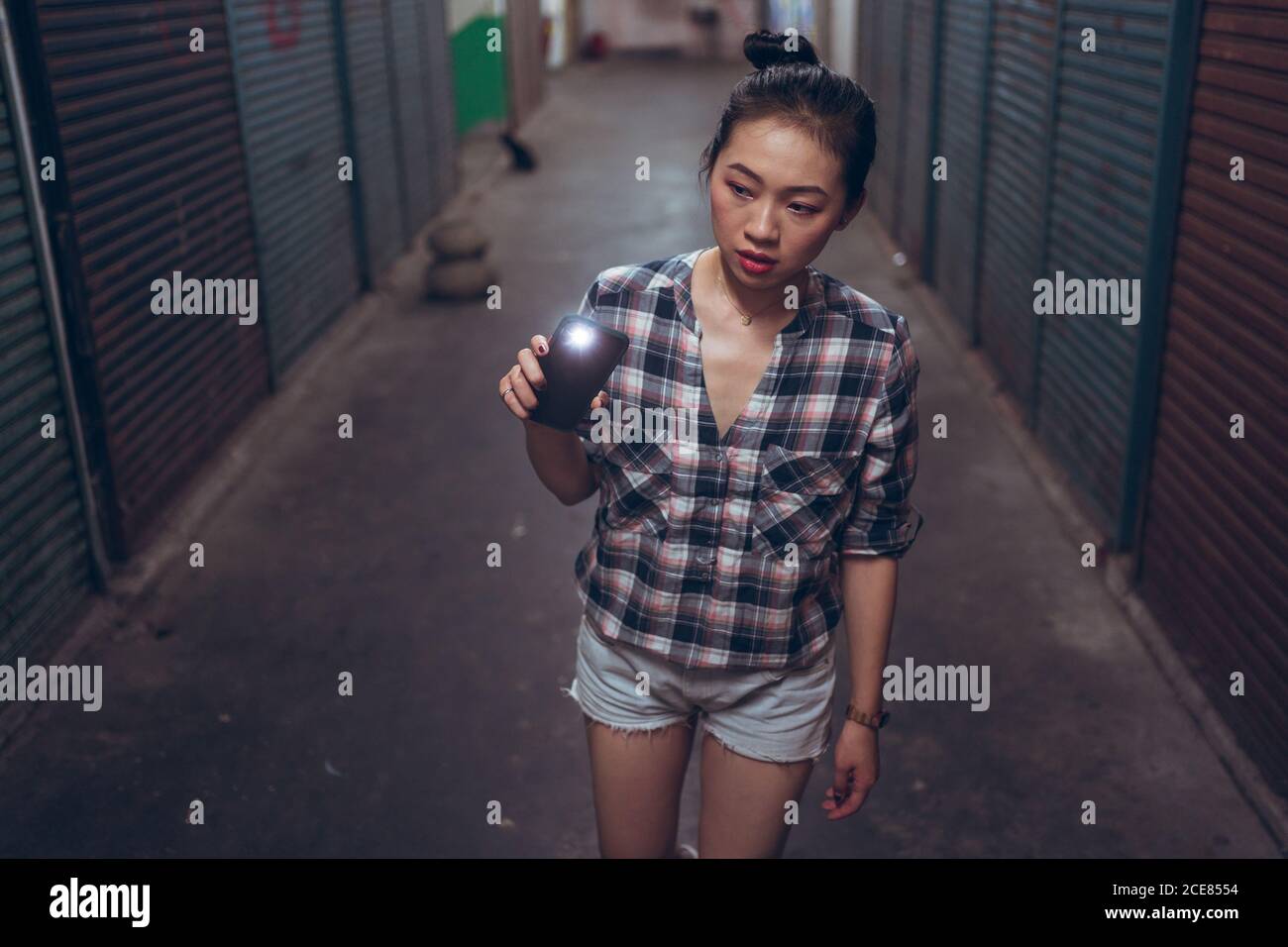Cautious young Asian female in casual clothes with glowing flashlight ...