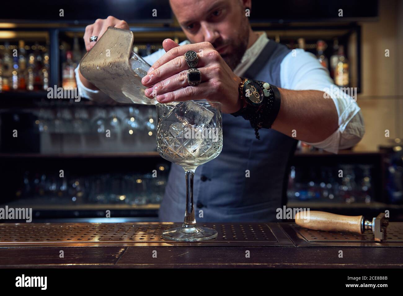 Focused bearded professional bartender pouring ice cubes into a cocktail in a glass while ...