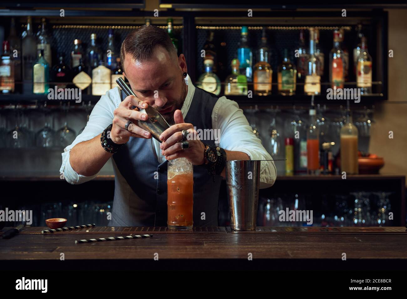 Bearded professional bartender pouring ice cubes into a cocktail in a glass while working in a ...