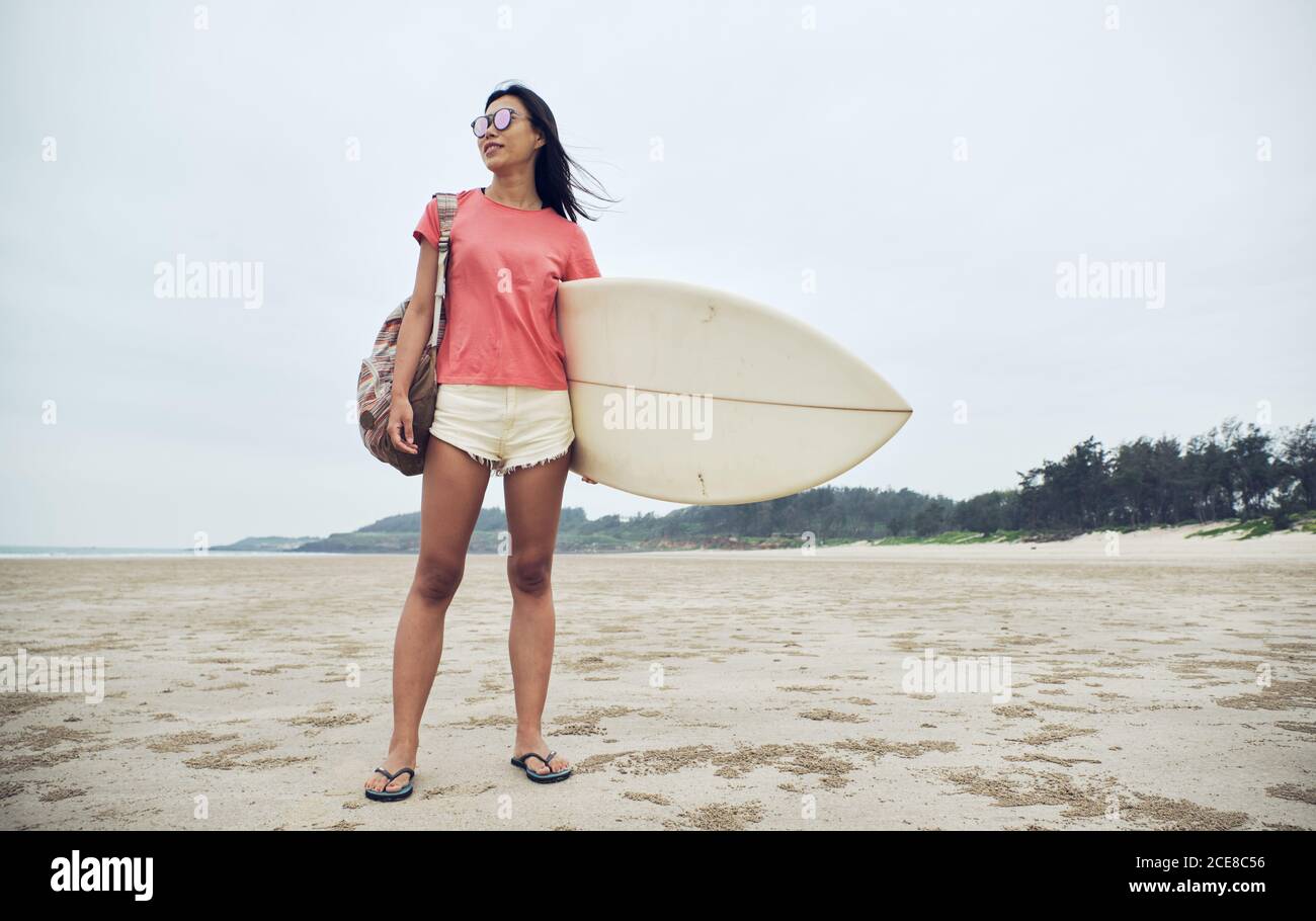 Full length young Asian female surfer in summer outfit walking on sandy ...