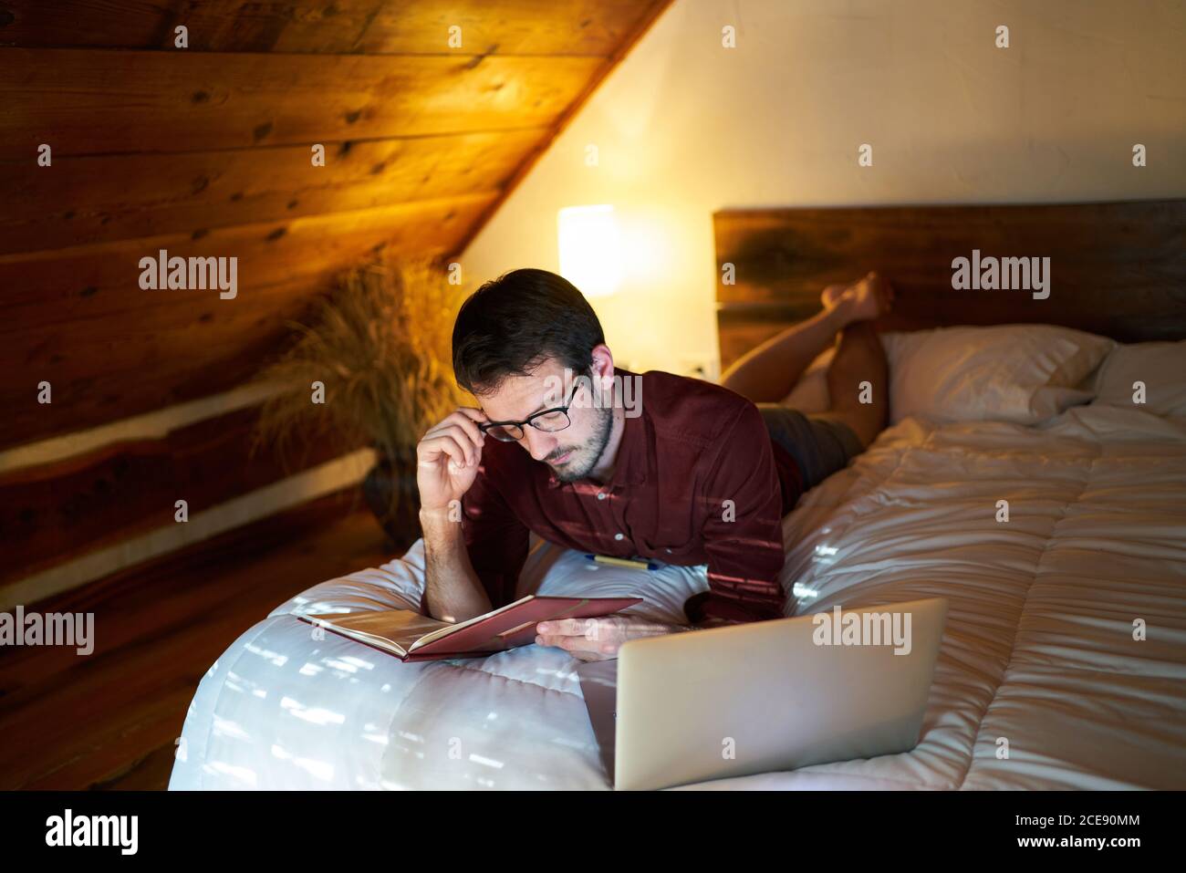 Busy male freelancer lying on bed with laptop in cozy bedroom and ...
