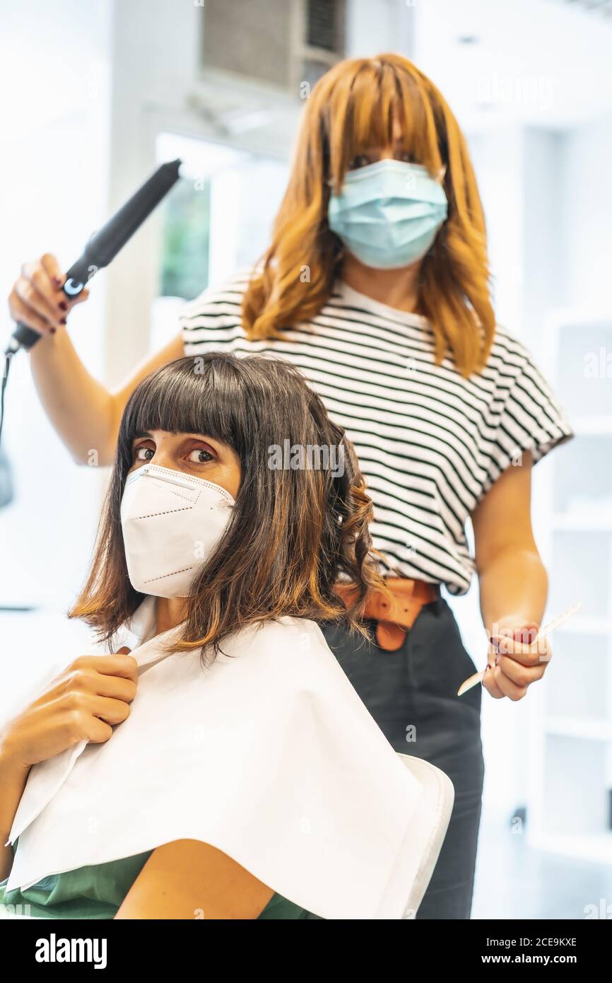 Female in a protective face mask getting a hairstyle with an iron hair ...