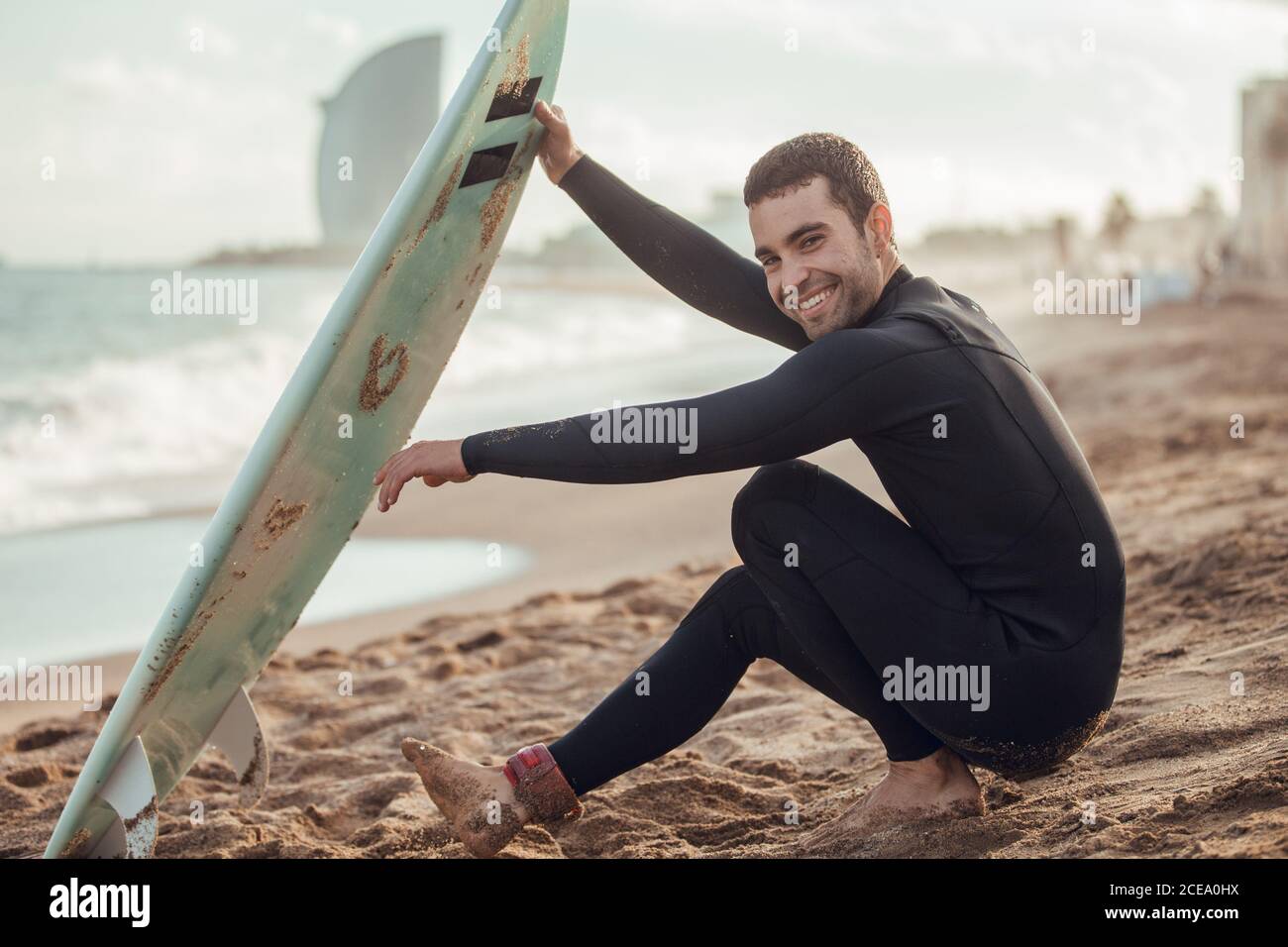Side view of man in black wetsuit sitting on sandy beach with surfboard ...