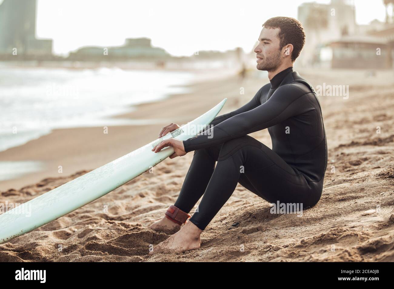 Side view of man in black wetsuit sitting on sandy beach with surfboard ...