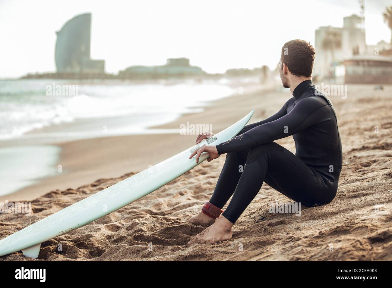 Side view of man in black wetsuit sitting on sandy beach with surfboard ...