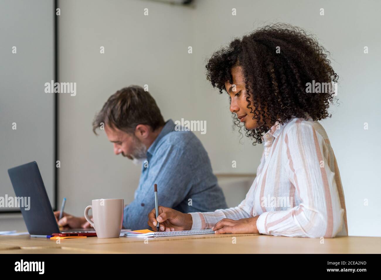 Worker sitting with colleague in office and writing in notebook Stock ...
