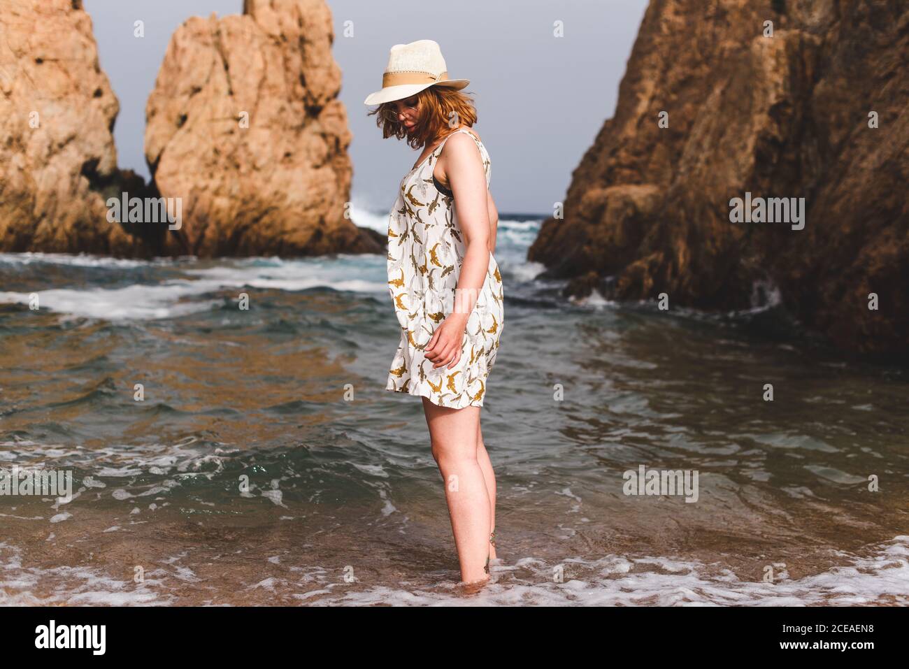 Pretty Woman in hat standing and relaxing in the ocean at big rocks ...