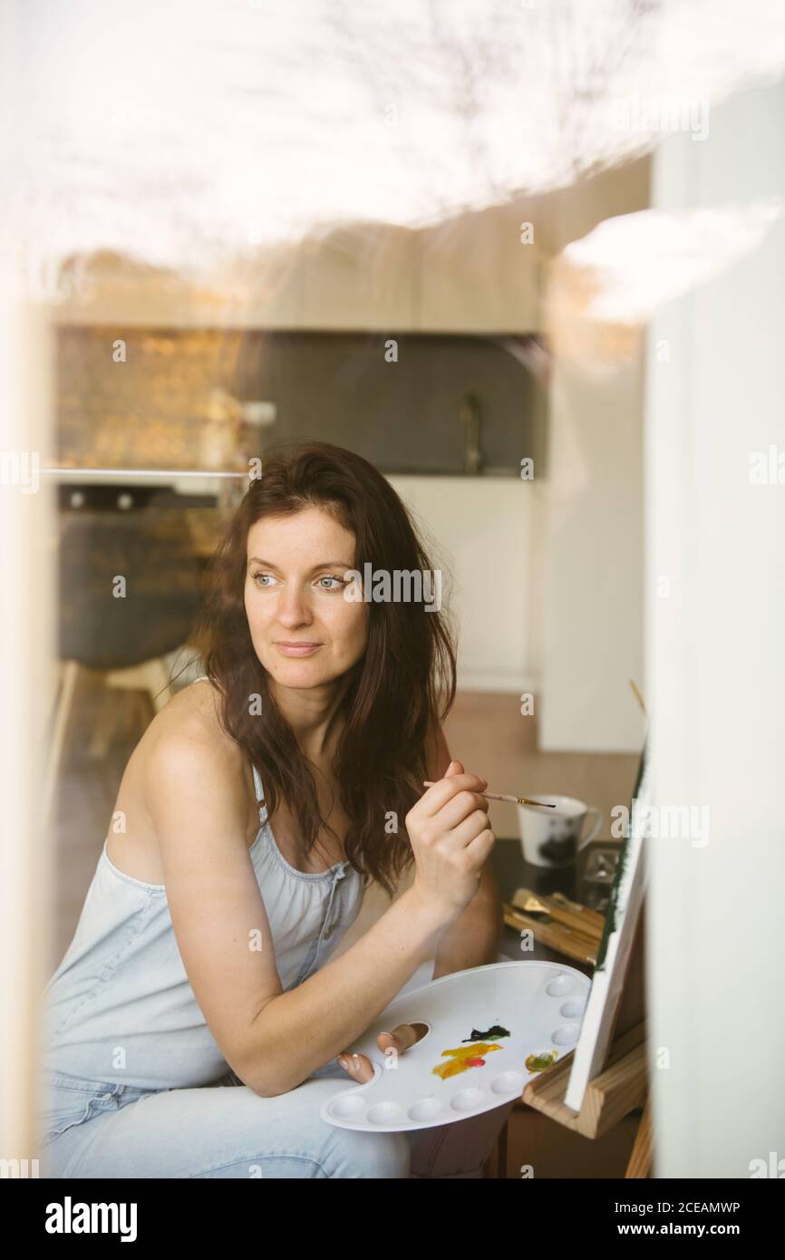Side view of beautiful Woman behind window glasses sitting at easel ...