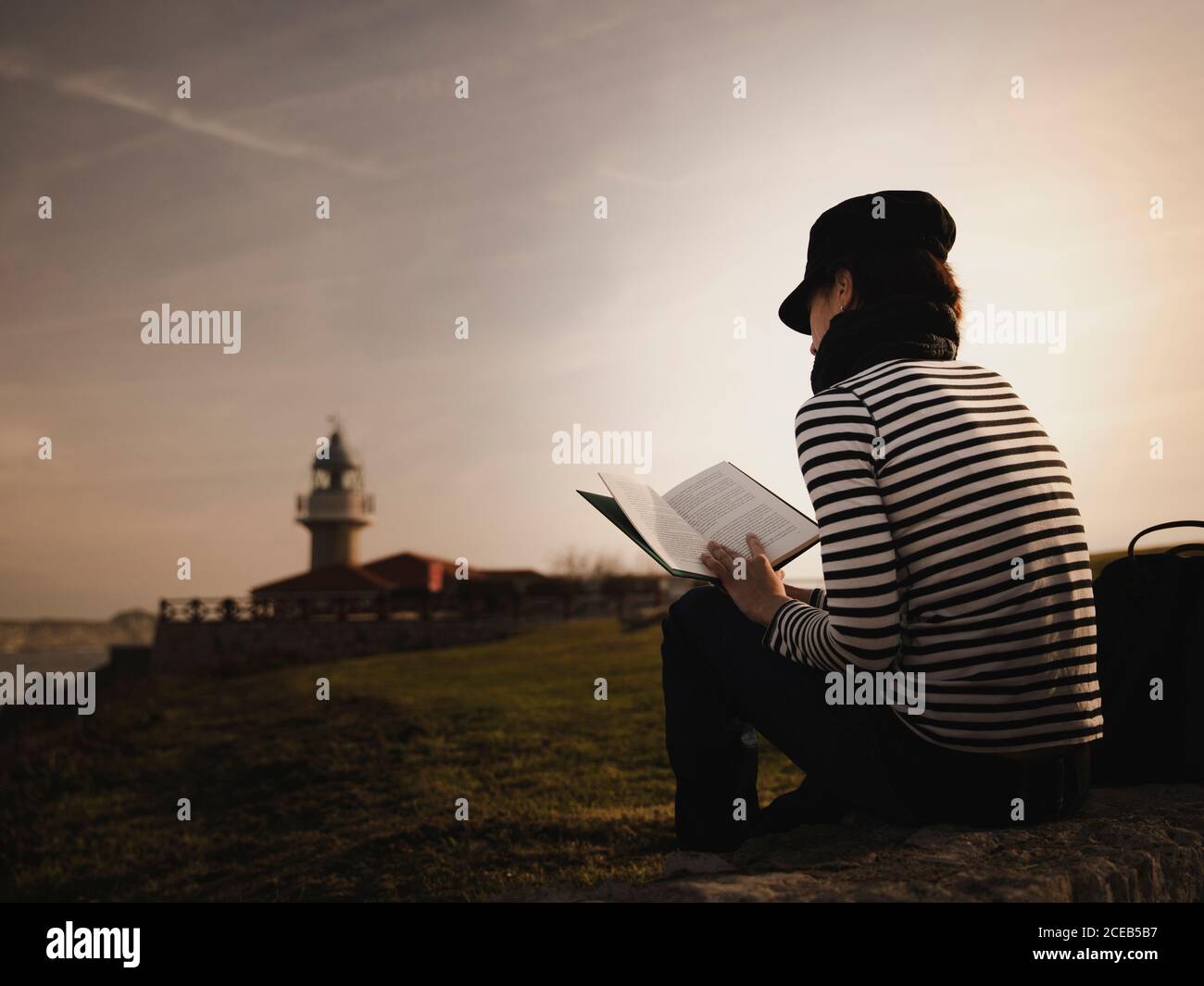 Back view of elegant lady in cap reading volume and sitting on meadow ...