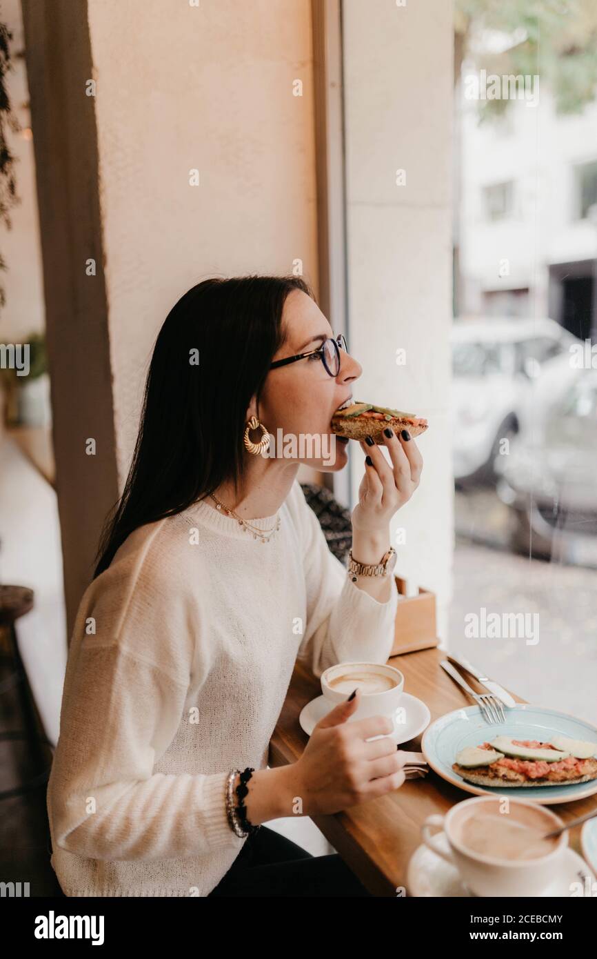 Side view of pretty young female in stylish outfit biting tasty ...