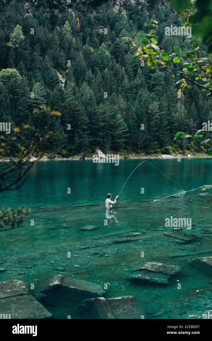 Back view of man standing in clean water of beautiful lake and fishing ...