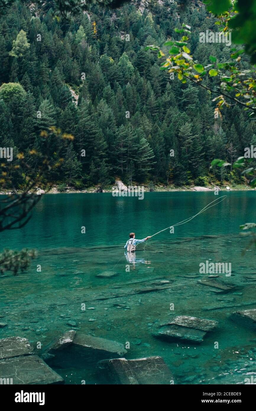 Back view of man standing in clean water of beautiful lake and fishing ...