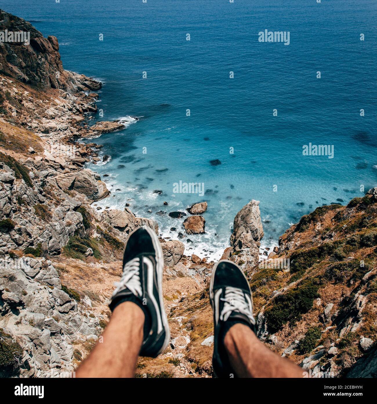 Crop legs of unrecognizable tourist sitting on cliff at blue ocean ...