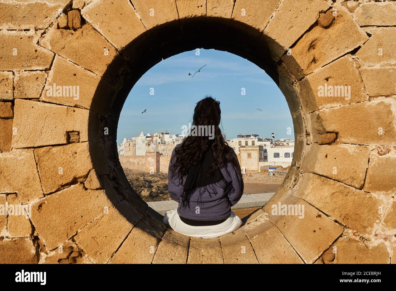 Back view of young brunette lady sitting on rock monument near old town ...