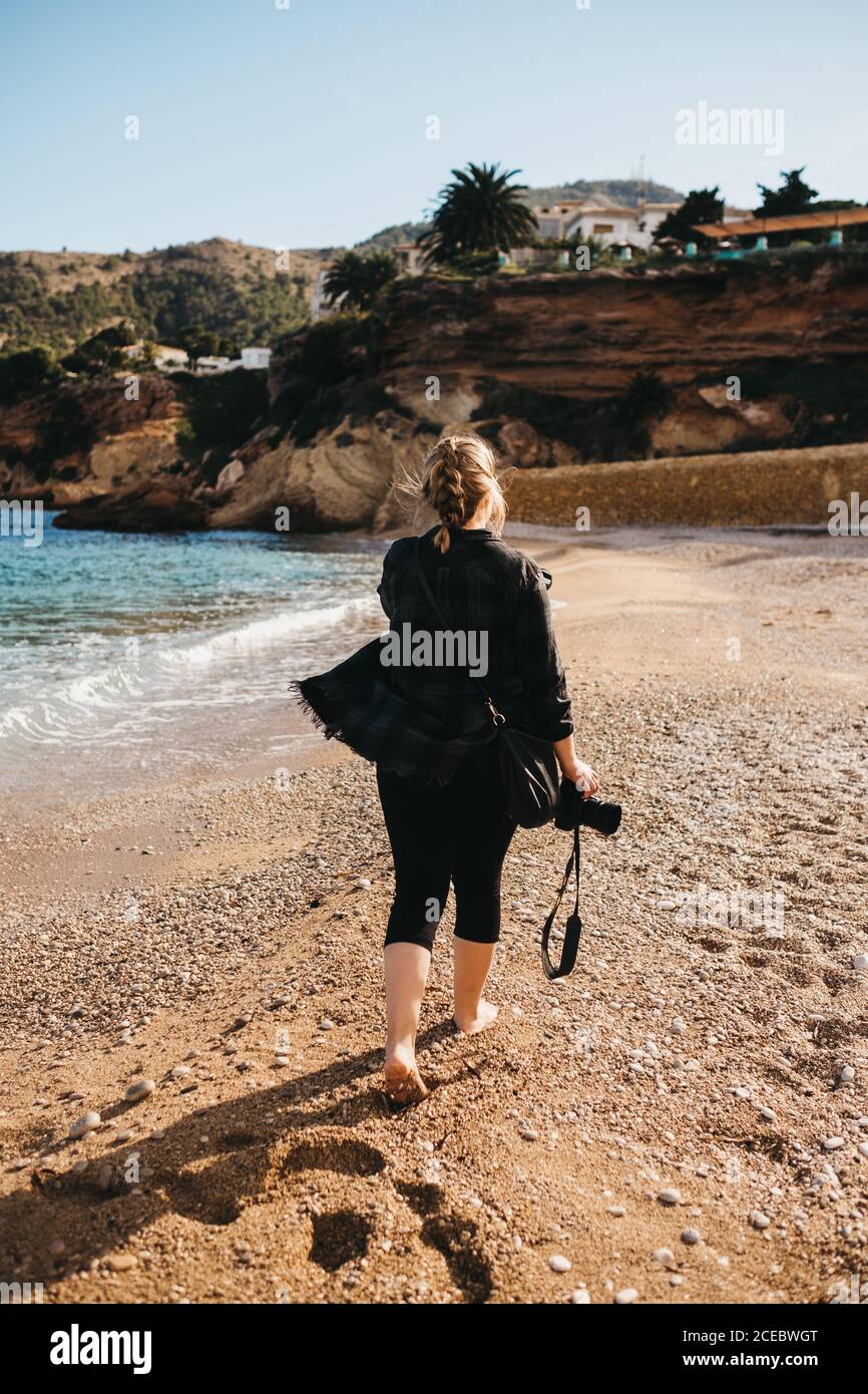 Back view of young barefoot lady with photo camera walking on sand near ...