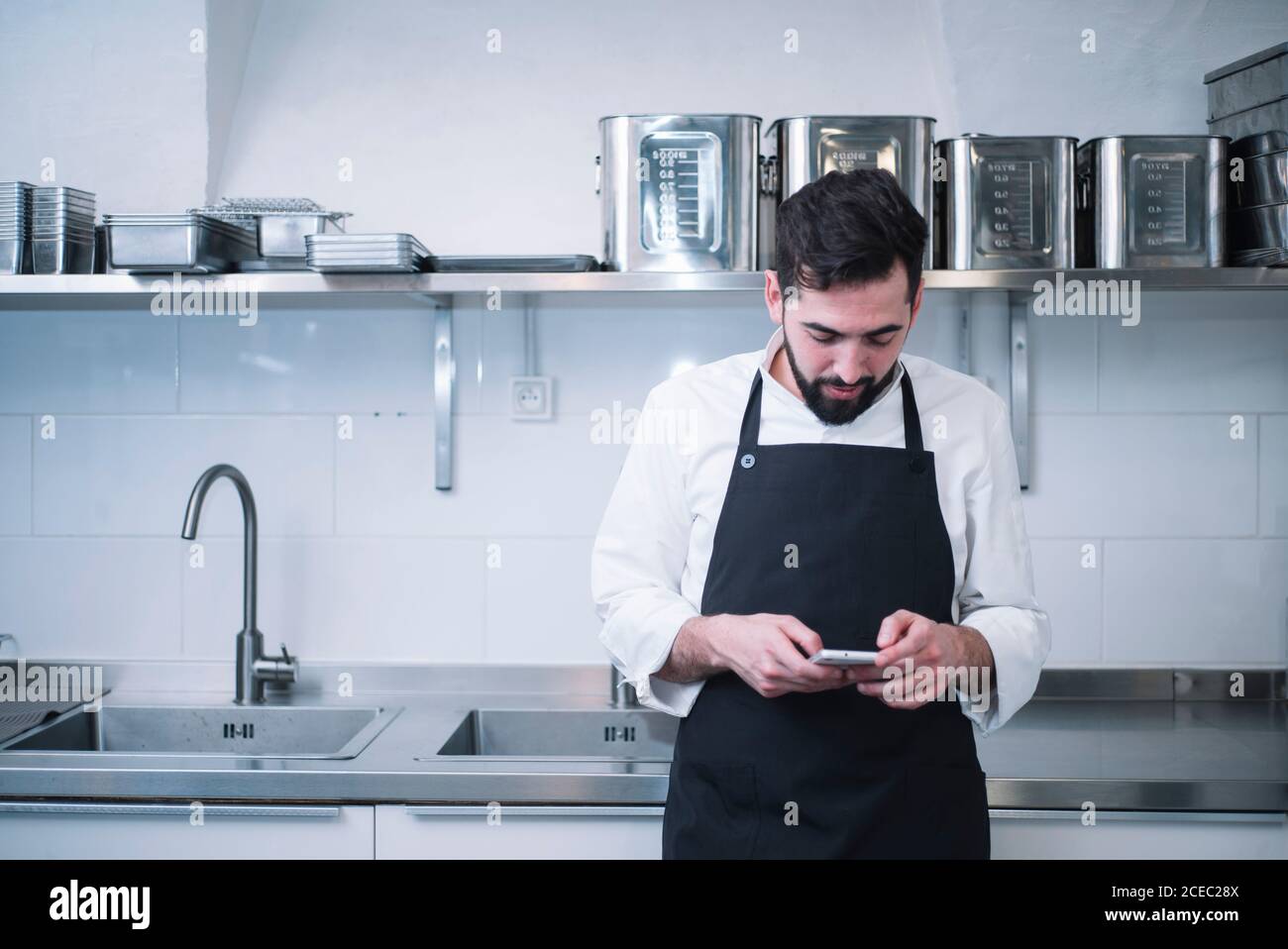 Side view of young man in cook uniform leaning on kitchen counter and ...