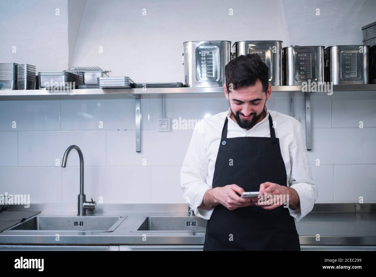 Side view of young man in cook uniform leaning on kitchen counter and ...