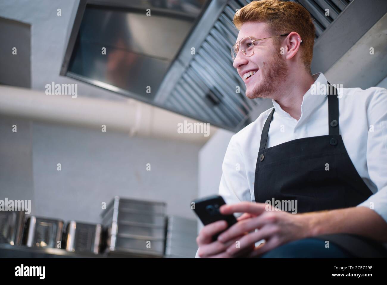 Side view of young man in cook uniform leaning on kitchen counter and ...