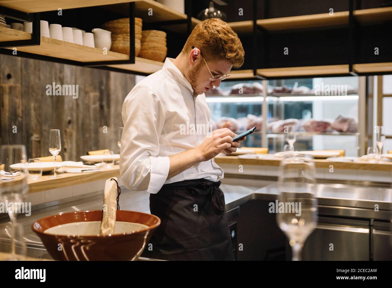 Side view of young man in cook uniform leaning on kitchen counter and ...