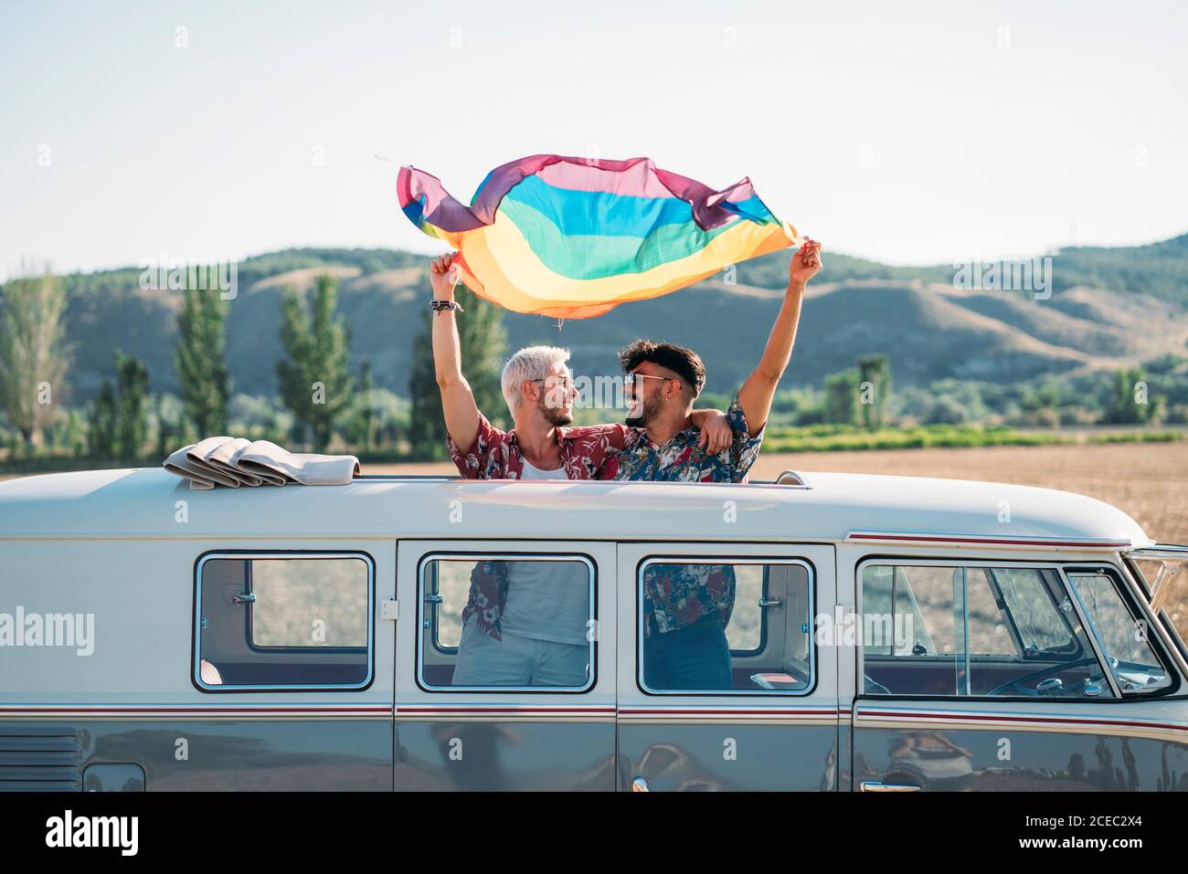 Two young men hugging and holding waving LGBT flag over heads while ...