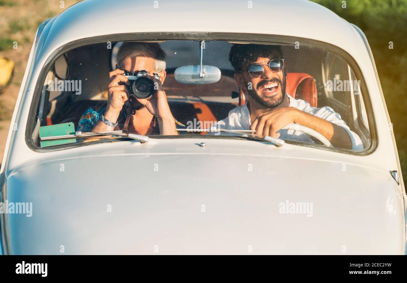 Two cheerful young guys with camera driving retro car and making ...