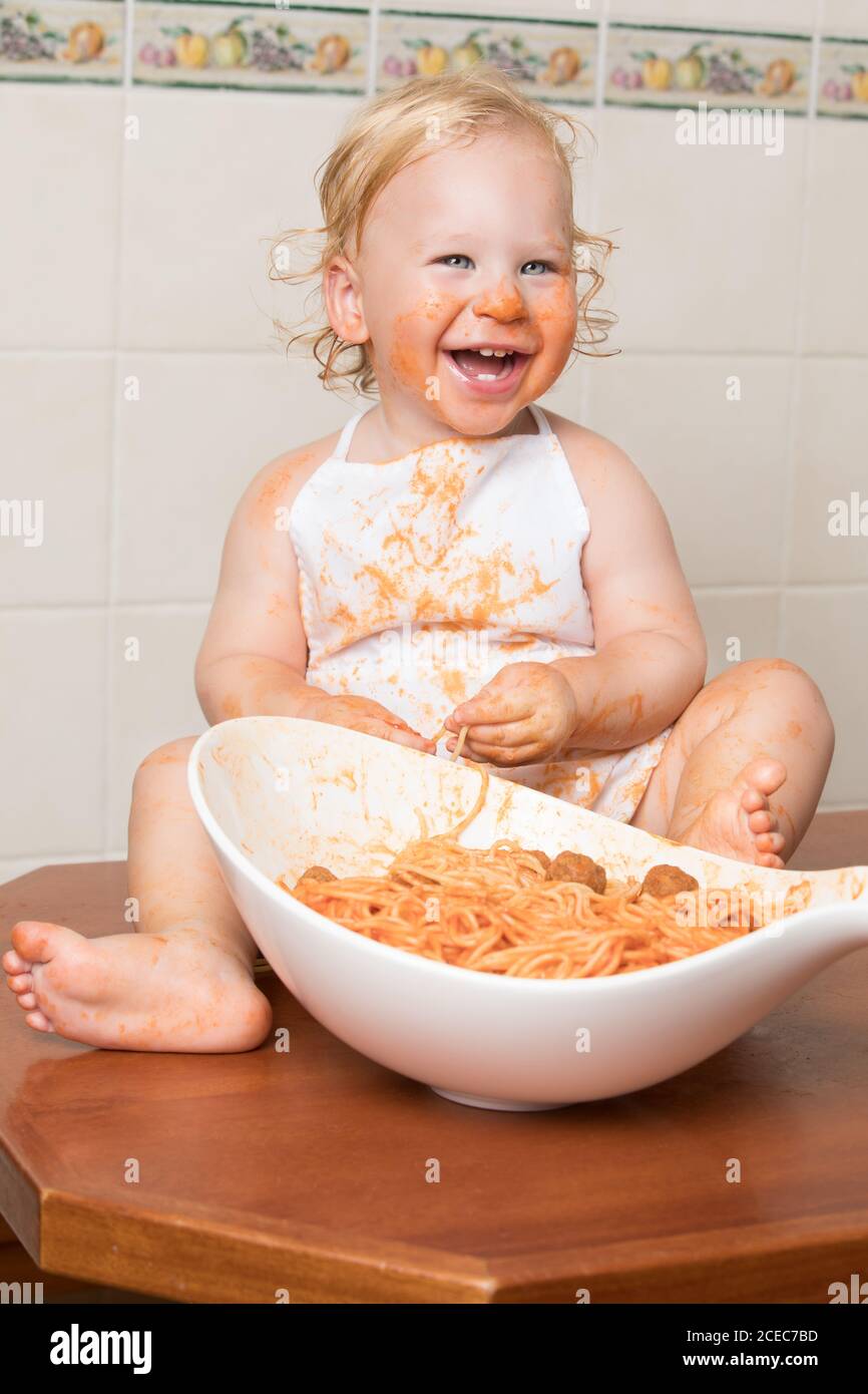 Cheerful little child sitting on tale and having fun while eating pasta ...