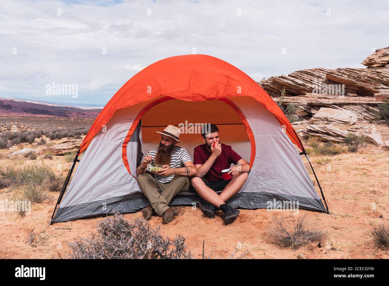 Two male friends smiling and sitting near tent together and eating ...