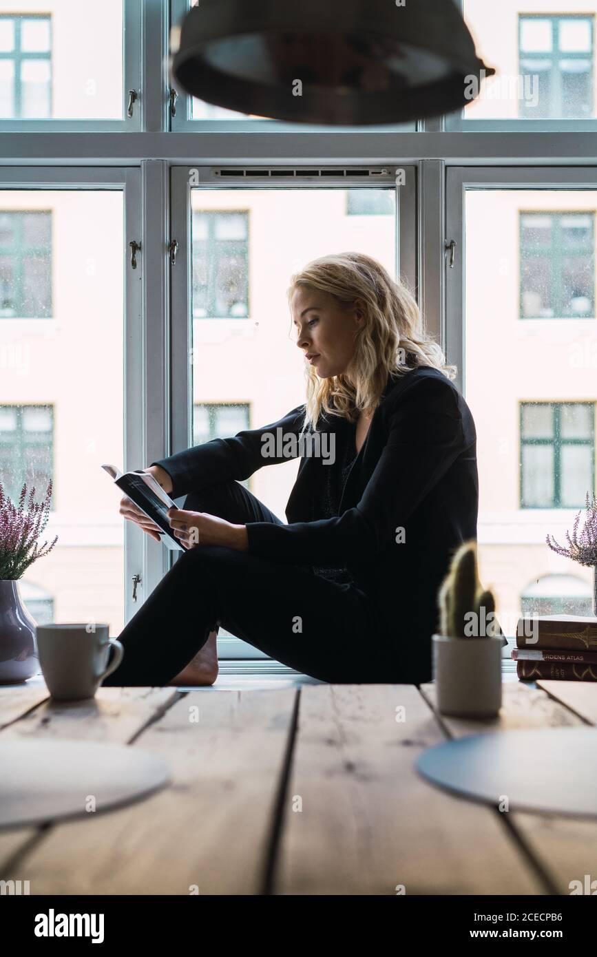 Young casual model immersed in book while lounging on window sill of ...