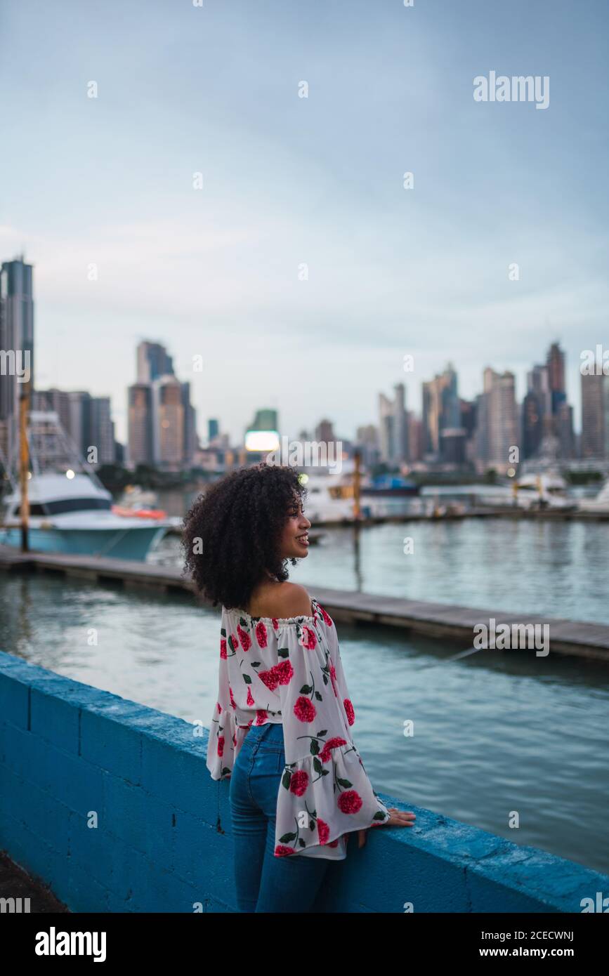 Side view of cheerful black Woman standing alone on colorful seafront ...