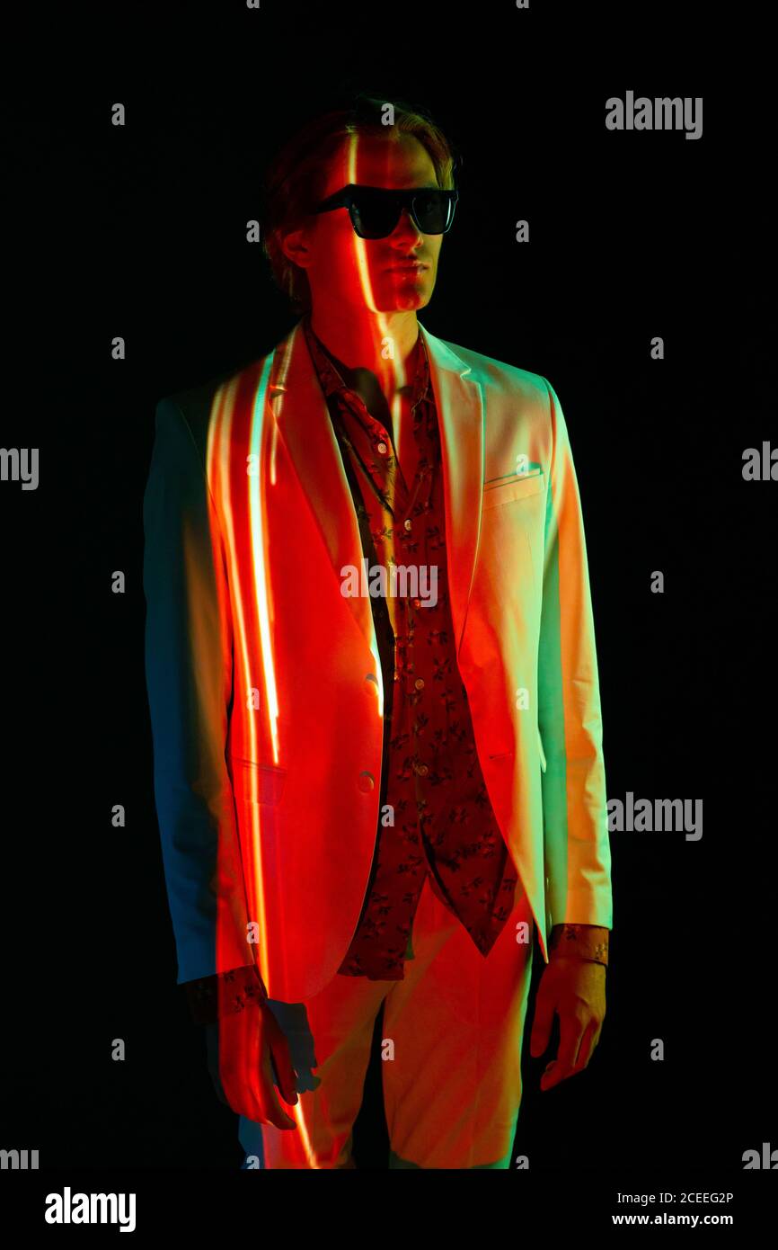 Androgynous male model in suit standing in relaxed pose under colorful illumination on black ...