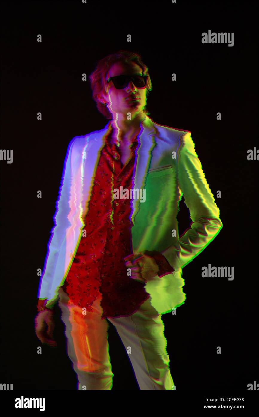 Androgynous male model in suit standing in relaxed pose under colorful illumination on black ...